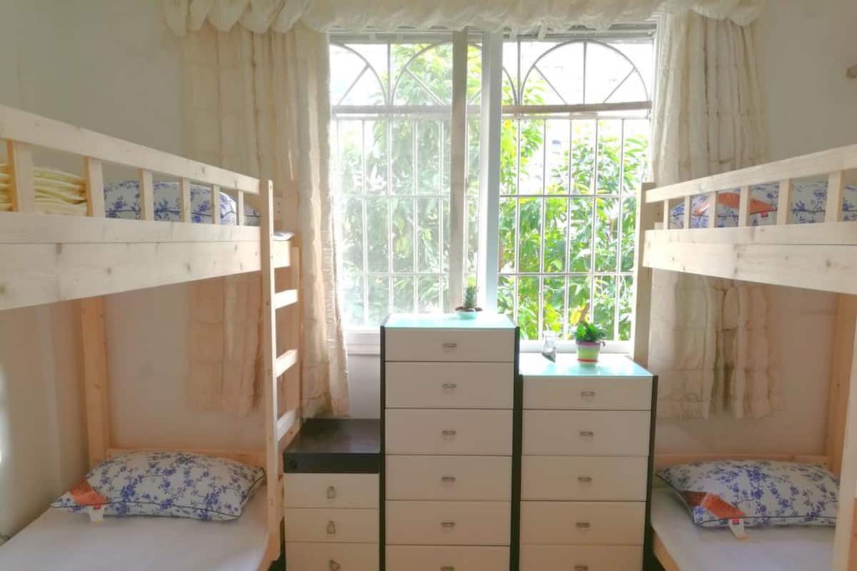 Quarto com beliches, uma escrivaninha central com gavetas e uma janela com cortinas.