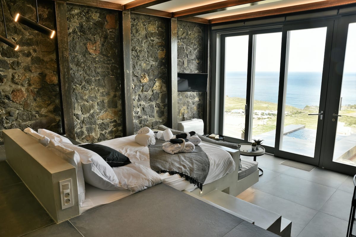 Quarto moderno com parede de pedra, uma cama com toalhas, uma mesinha e vista para o mar.