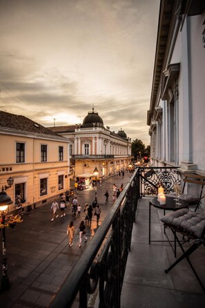 Varanda com vista para uma rua movimentada, prédios históricos, um mercado e pedestres.