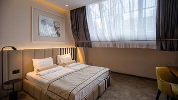 Quarto de hotel moderno com uma cama grande, uma mesinha de cabeceira, uma cadeira e uma janela com cortinas.