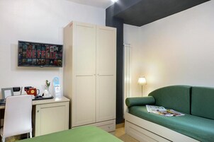Quarto com cama, escrivaninha com computador, armário, sofá verde e TV na parede exibindo Netflix.