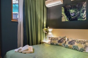 Uma cama bem arrumada com um cobre-leito verde e almofadas estampadas com folhas tropicais, um vaso com flores em uma mesa de cabeceira e um ar condicionado fixado na parede.