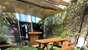 Um pátio externo com mesas de piquenique de madeira, grama sintética verde, plantas em vasos e uma área coberta com piso de concreto.