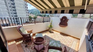 Varanda com mobiliário de vime, chão estampado e vista para edifícios ao fundo.
