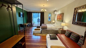 Quarto de hotel com cama, sofá, escrivaninha, cadeira, banqueta, lustre, vitral e pintura.