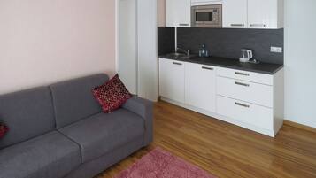 Um espaço compacto para viver com sofá cinzento, tapete rosa, cozinha americana com armários brancos, micro-ondas e máquina de café.