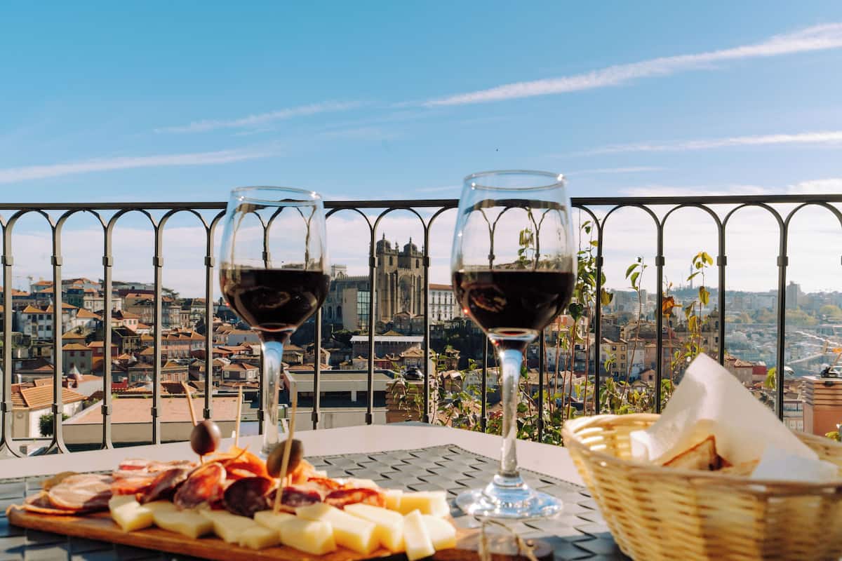 Uma varanda com vista para a cidade, uma mesa com tábua de frios e duas taças de vinho tinto.