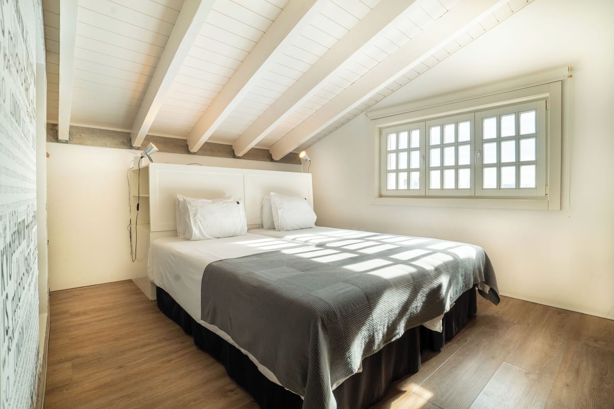 Quarto com cama, piso de madeira, paredes brancas, janela e teto inclinado.