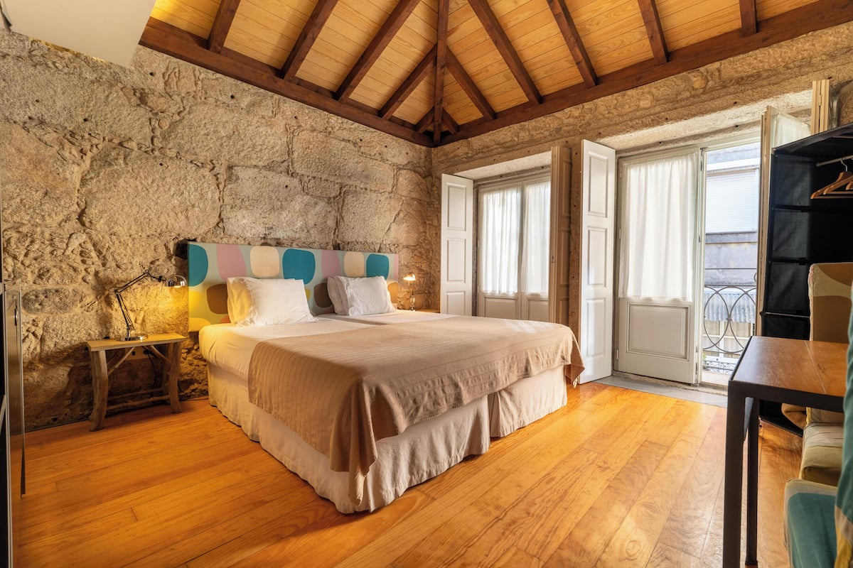 Quarto com parede de pedra, vigas de madeira no teto, cama com cabeceira colorida e chão de madeira.