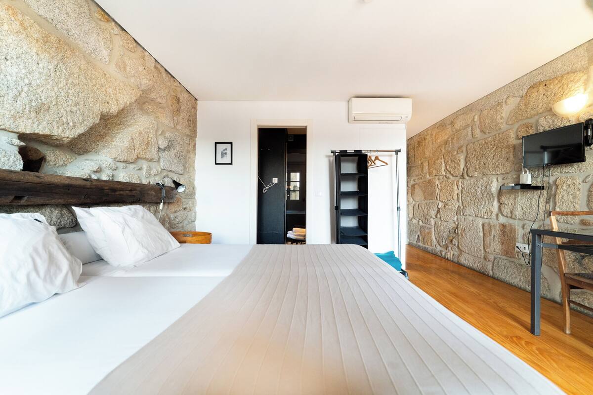 Quarto com parede de pedra, cama com roupa de cama branca, piso de madeira e televisão fixada na parede.