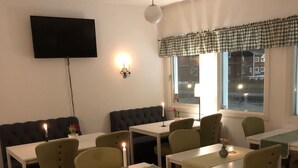 Uma sala de jantar com mesas e cadeiras, uma televisão na parede e uma janela com vista para o exterior.