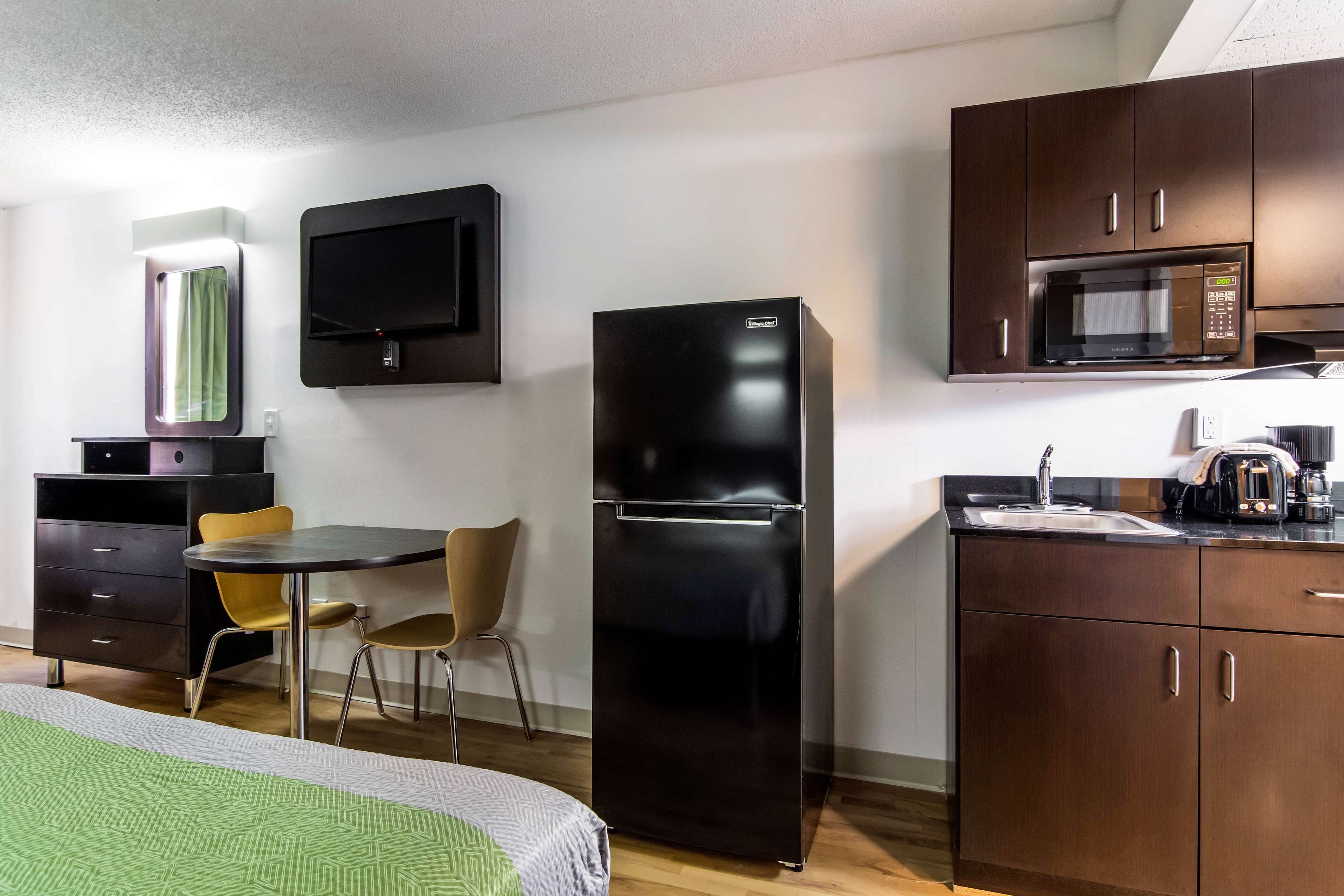 Quarto de hotel compacto com uma cama, uma pequena mesa de jantar, uma cadeira, um frigorífico preto, um micro-ondas e uma TV na parede.