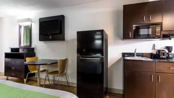Quarto de hotel compacto com uma cama, uma pequena mesa de jantar, uma cadeira, um frigorífico preto, um micro-ondas e uma TV na parede.