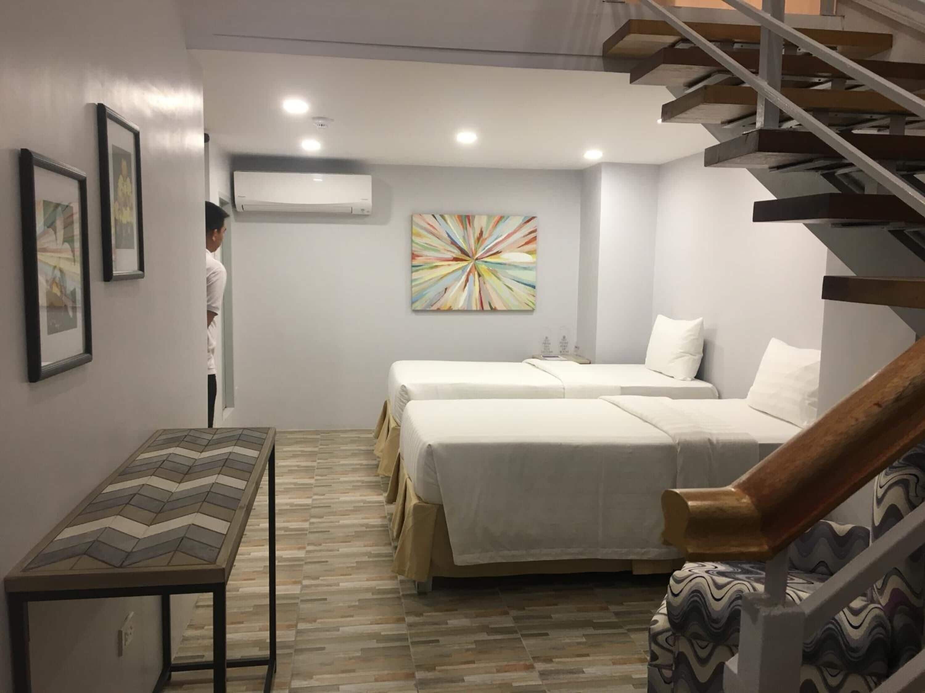 Quarto de hotel com duas camas, um banco, uma escada e um aparelho de ar condicionado.
