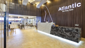 Uma área de recepção de hotel com um balcão de mármore, um visor digital e uma área de estar com mesas e cadeiras.