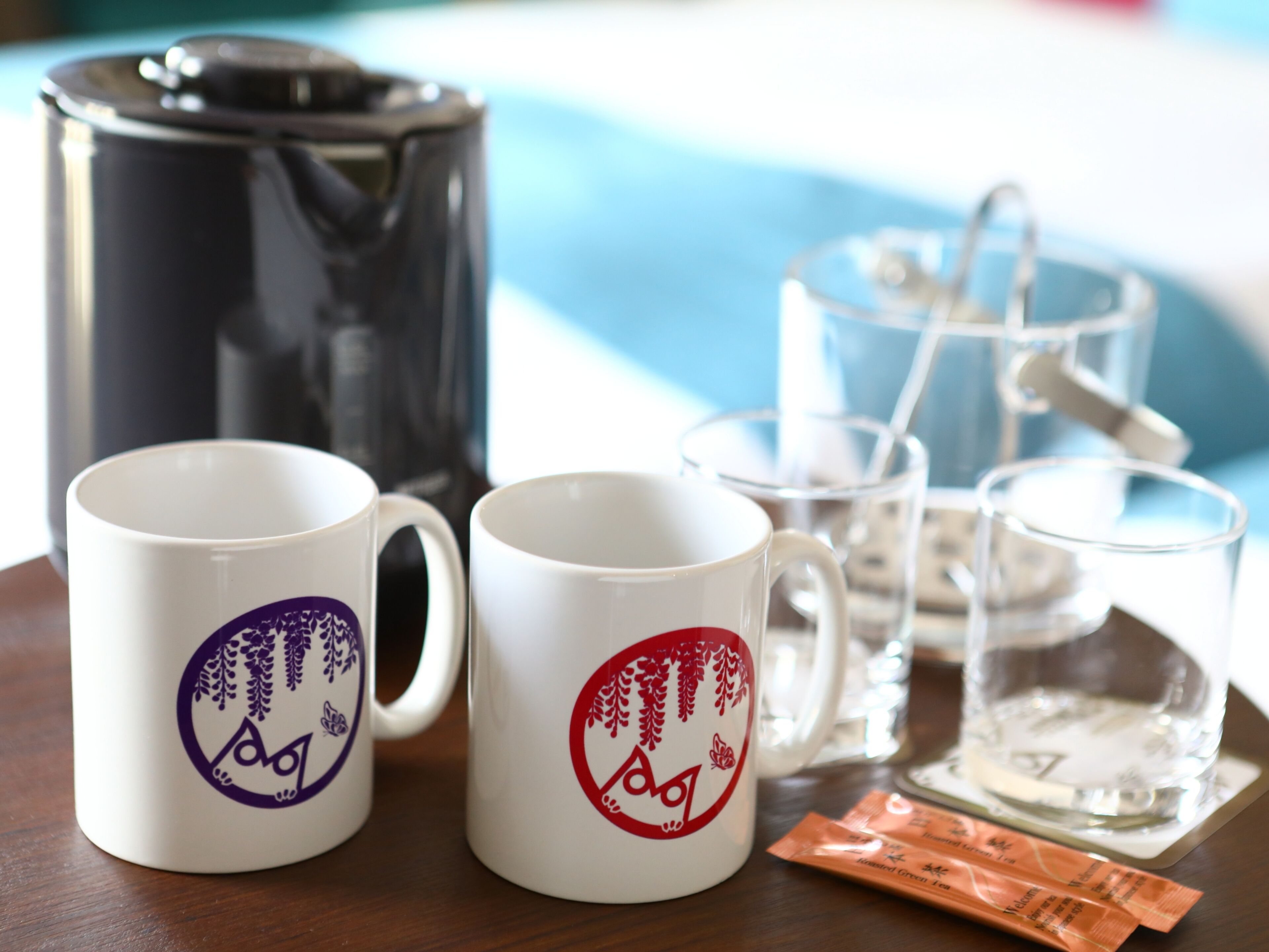 Duas canecas brancas com logotipos, uma cafeteira preta, uma chávena de vidro e dois sachês de chá sobre uma superfície de madeira.