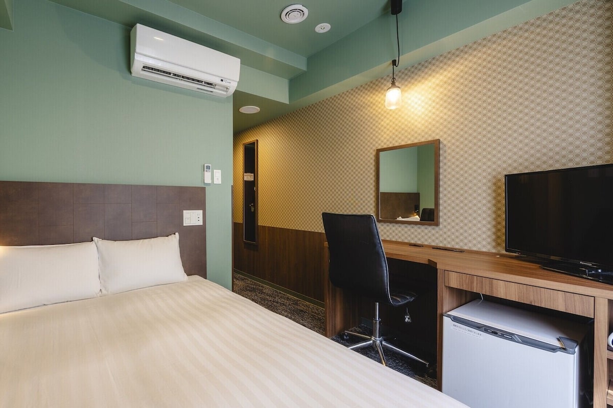 Quarto de hotel com cama, escrivaninha, cadeira, televisão, ar condicionado e espelho.