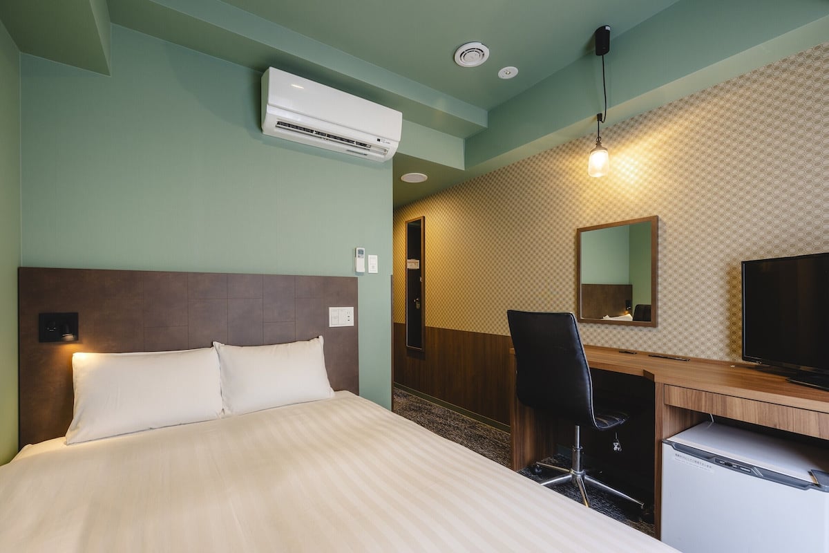 Quarto de hotel com cama, escrivaninha, cadeira, televisão e ar condicionado.