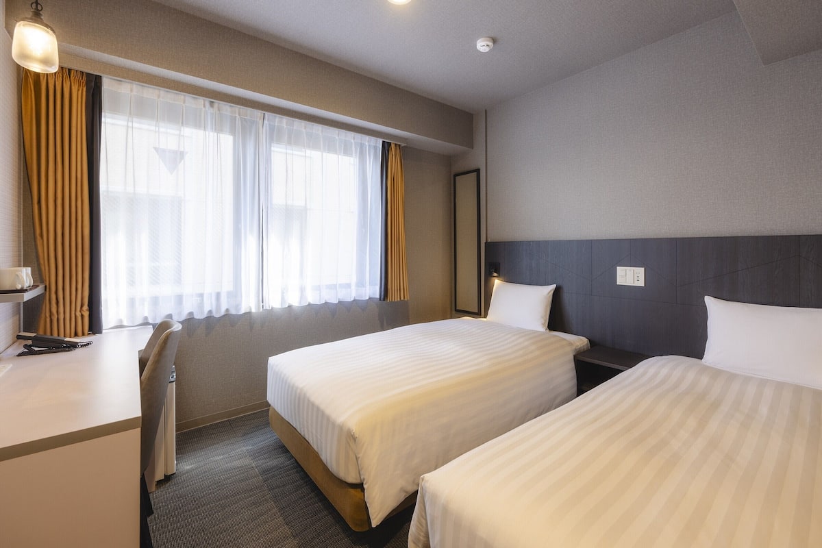 Quarto de hotel com duas camas, uma mesa, uma cadeira e uma janela grande com cortinas.