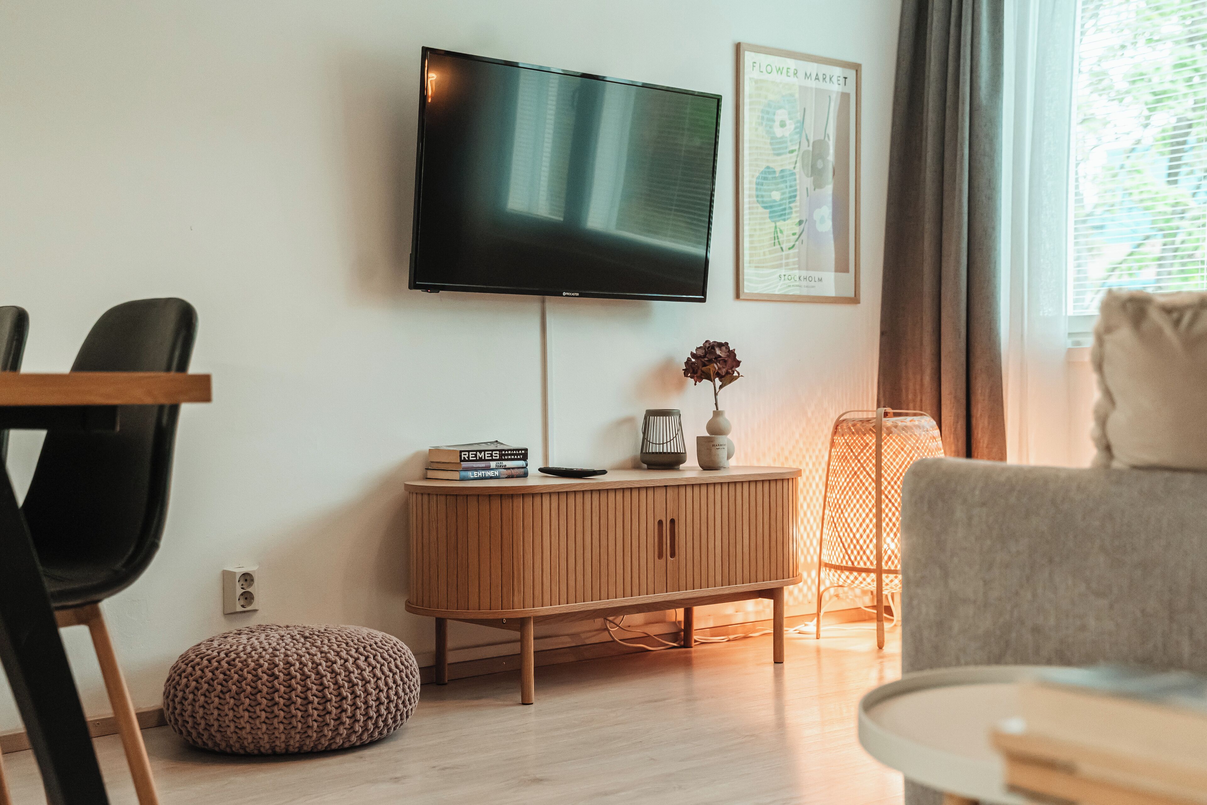 Uma sala de estar moderna com uma TV de tela plana na parede, um armário de madeira, um banquinho redondo e uma moldura com um quadro na parede.