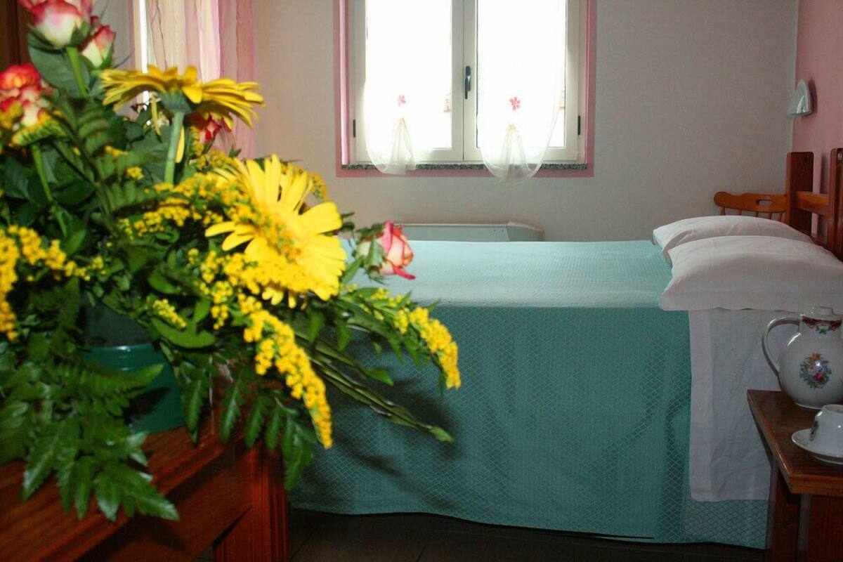 Quarto com cama, janela com cortinas leves, um vaso de flores e uma mesa de cabeceira com bule.