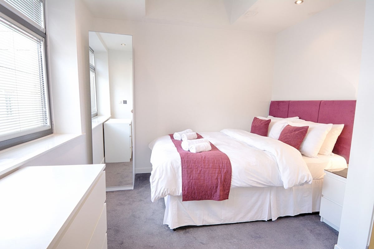 Quarto com cama, roupa de cama branca, cabeceira rosa e janela com persianas.