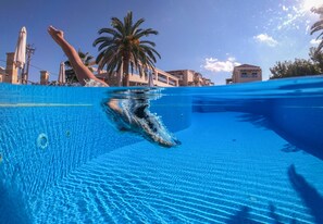 Vista subaquática de uma piscina com água azul cristalina, palmeiras e edifícios ao fundo.