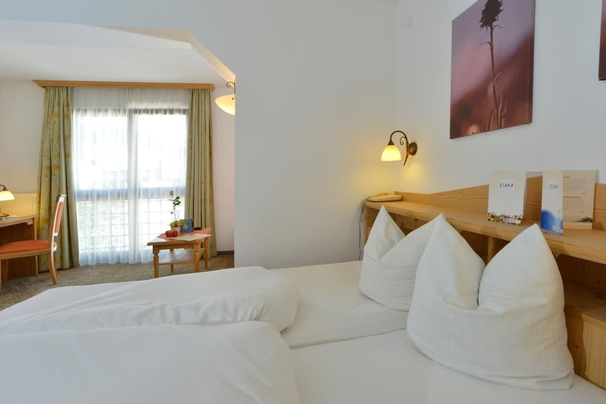 Quarto de hotel com cama, escrivaninha, cadeira, luminária, janela com cortinas e telefone fixado na parede.