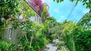 Um caminho de pedra estreito, ladeado por vegetação exuberante e plantas floridas, conduz a um edifício com fachada branca e telhado de telhas vermelhas.