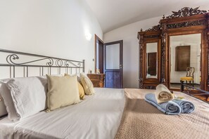 Cama bem arrumada com travesseiros, um espelho grande e ornamentado, e uma porta de madeira em um quarto com paredes brancas.