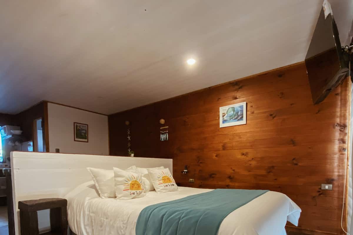 Um quarto com cabeceira de madeira, cama com roupa de cama branca e manta verde-água, uma mesinha de cabeceira e uma TV fixada na parede.