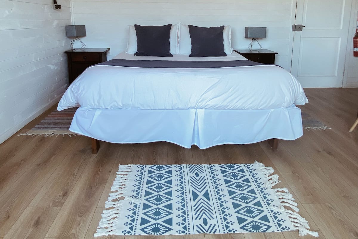 Quarto com cama, duas mesas de cabeceira, luminária na parede, porta e tapete estampado.