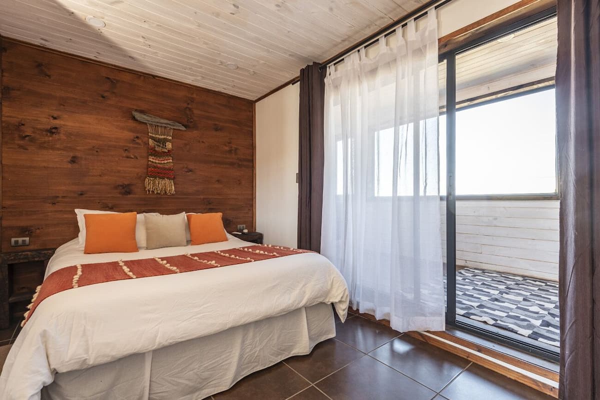 Quarto com parede de madeira, cama com roupa de cama branca e laranja, e uma janela grande com cortinas leves.