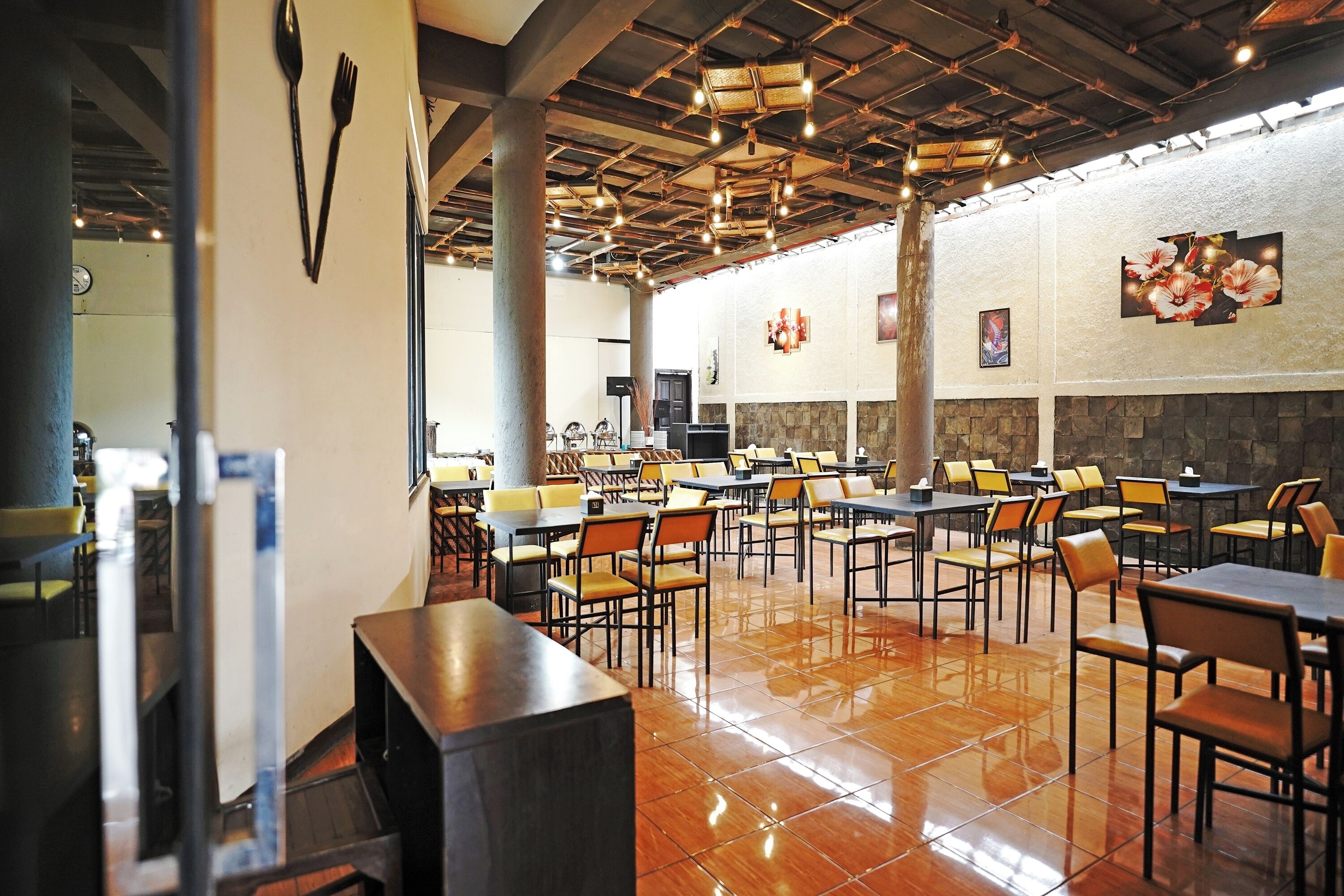 Uma ampla sala de jantar com várias mesas e cadeiras, teto alto com vigas aparentes e parede de pedra com obras de arte.