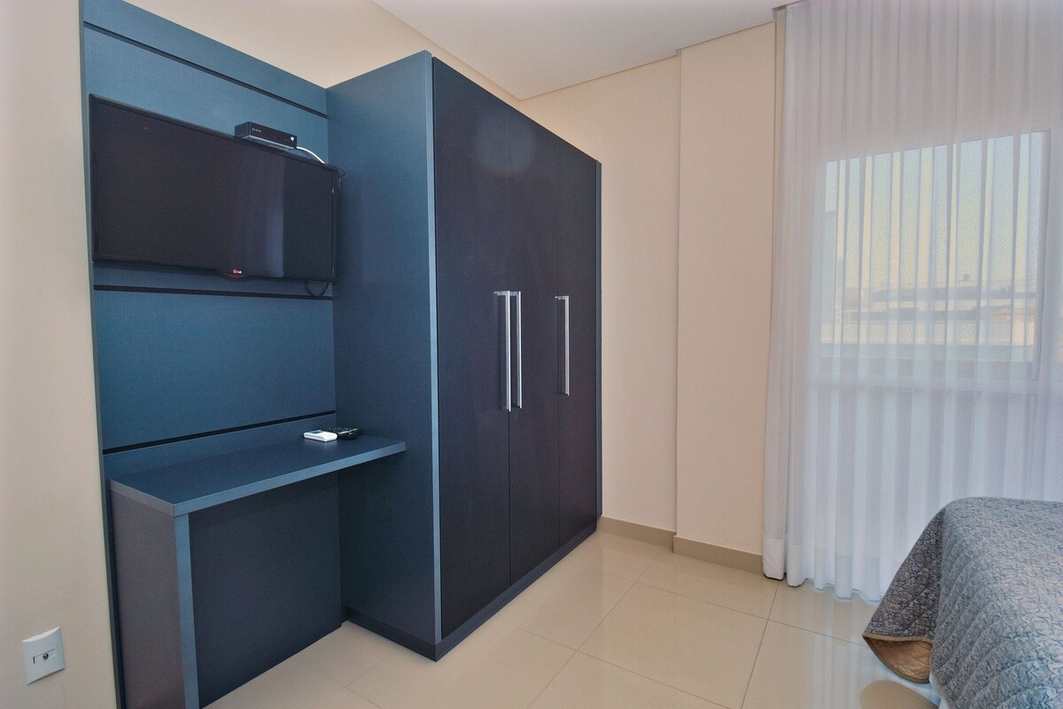 Quarto com um guarda-roupa azul escuro, televisão de tela plana na parede e uma cama com colcha estampada.