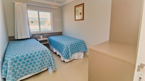 Quarto pequeno e limpo com duas camas de solteiro, uma janela com cortinas, uma mesinha e um quadro na parede.