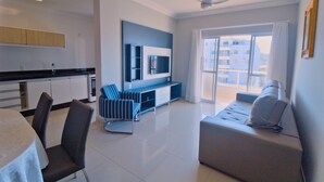 Uma sala de estar moderna com um sofá, uma mesa de jantar com cadeiras, uma TV fixada na parede e uma varanda com vista para edifícios.