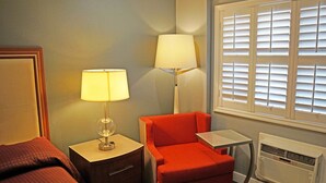 Quarto de hotel com cama, poltrona vermelha, mesinha, duas luminárias e janela com persianas.