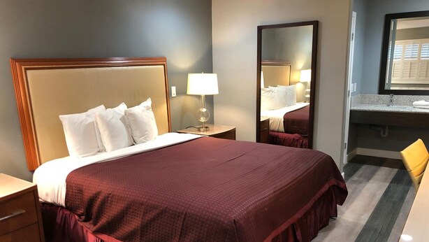 Quarto de hotel com uma cama grande, uma mesa de cabeceira, uma luminária, um espelho, um banheiro com pia e espelho, e uma cadeira.