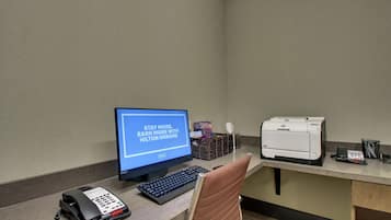 Um espaço de trabalho em hotel com computador, telefone, impressora e um painel promocional.