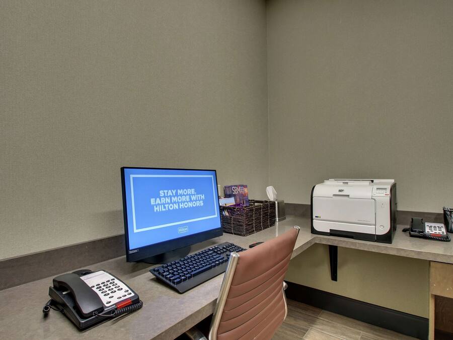 Um espaço de trabalho em hotel com computador, telefone, impressora e um painel promocional.