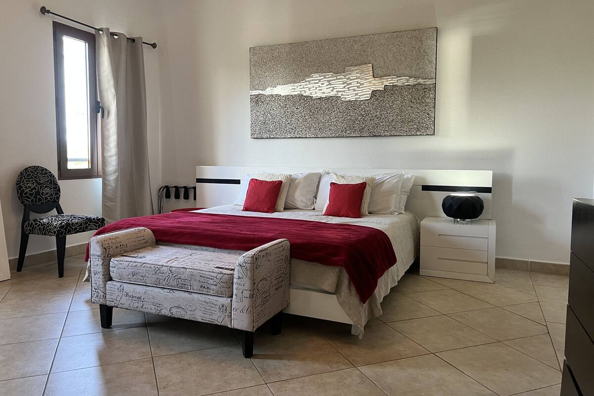 Quarto com cama, cadeira, mesa de cabeceira, luminária e uma obra de arte na parede.