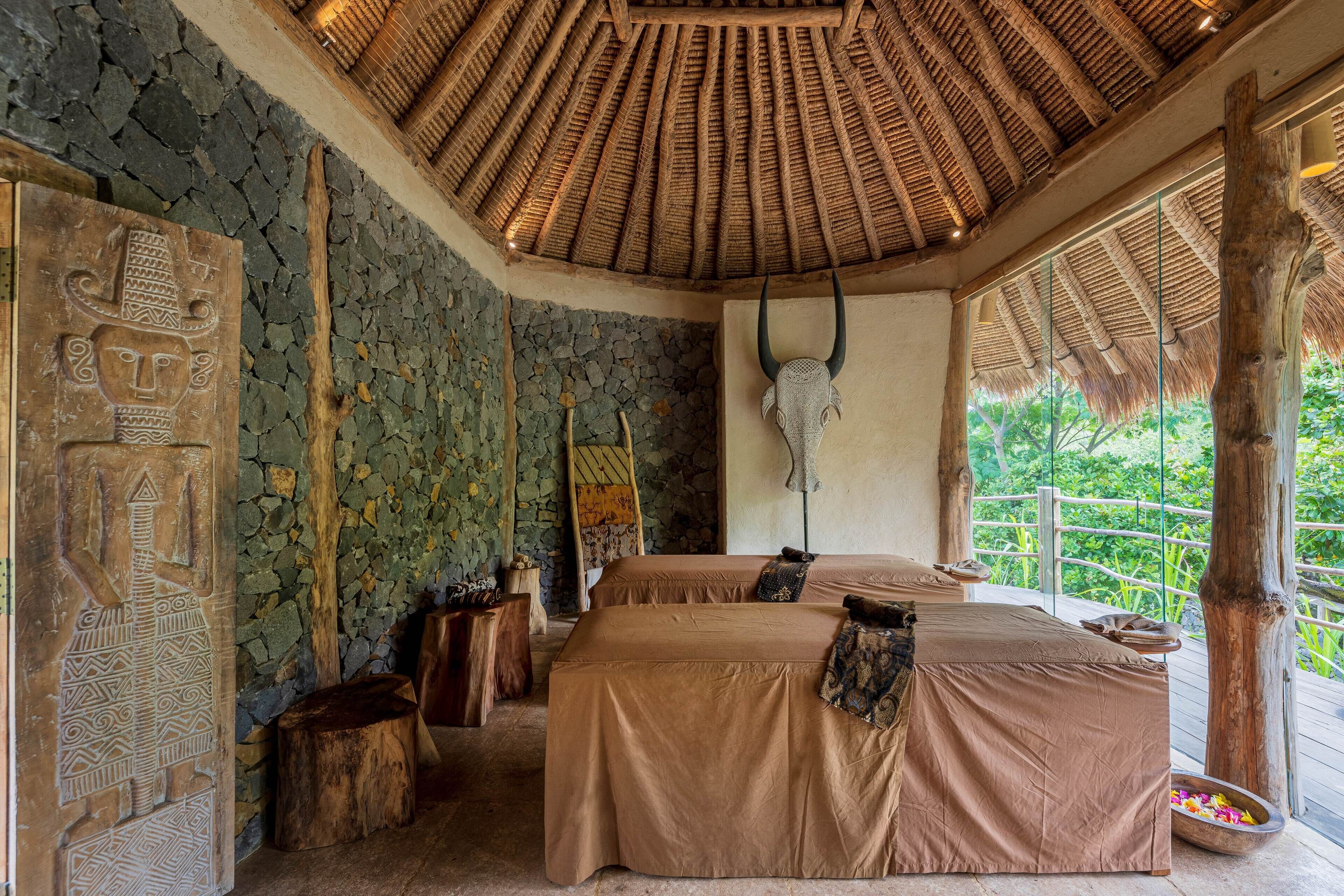 Quarto com duas camas, parede de pedra e teto de palha.