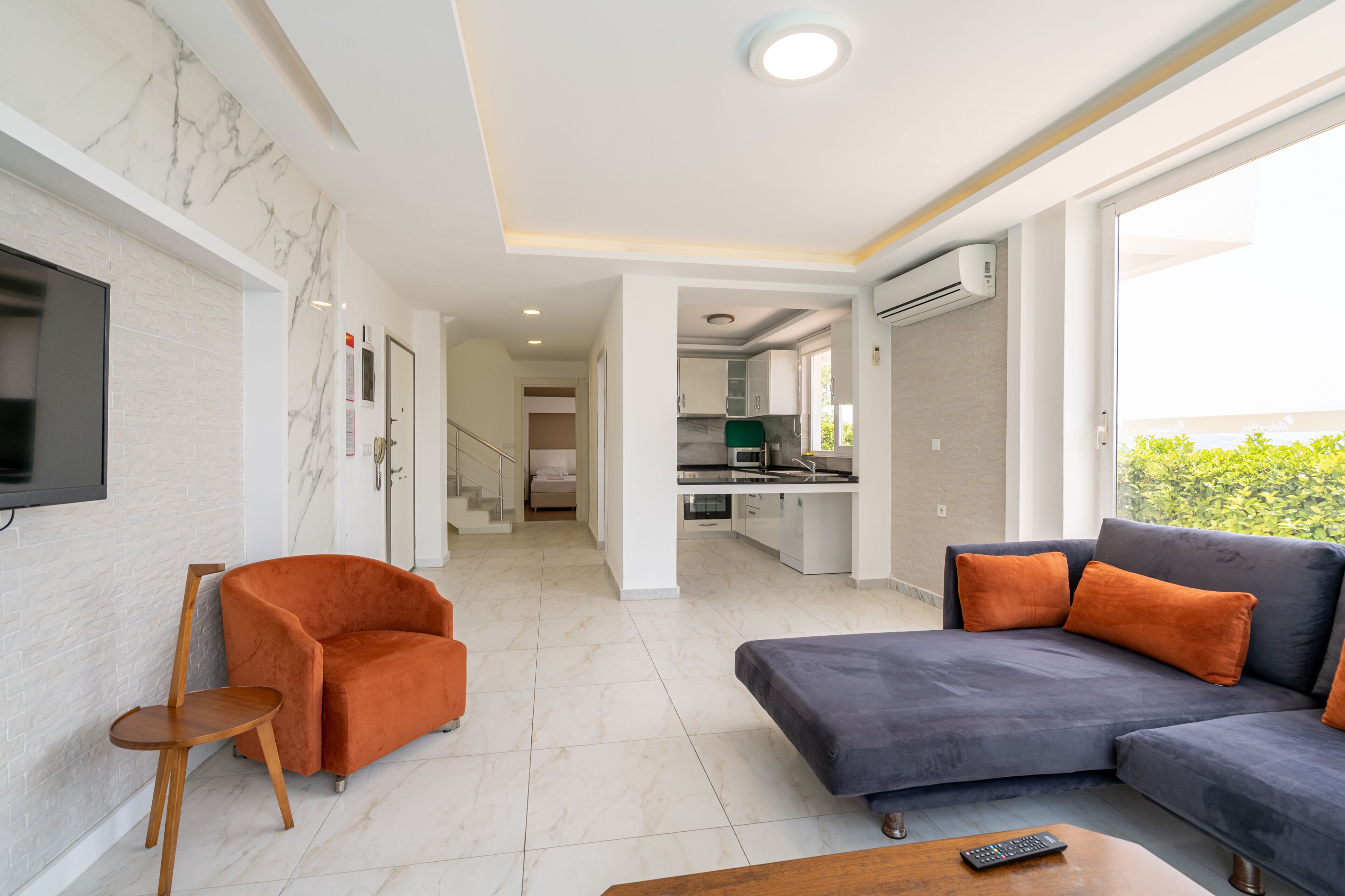 Uma sala de estar moderna com um sofá, poltrona e mesa de centro. Uma cozinha é visível ao fundo.