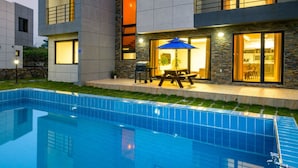 Uma casa moderna de dois andares com piscina, janelas amplas e um pátio com mobiliário de exterior.