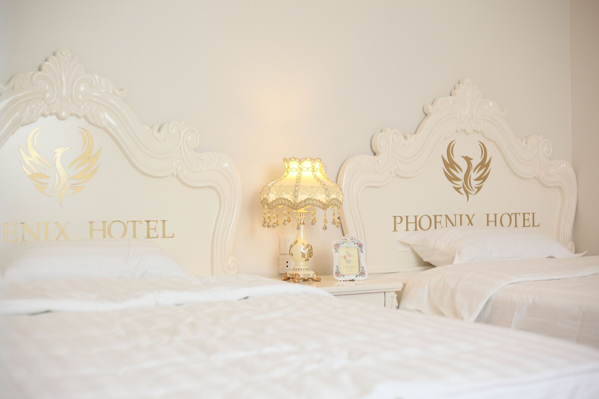 Quarto de hotel com duas camas, cabeceiras decoradas, mesa de cabeceira com abajur e moldura ornamental.