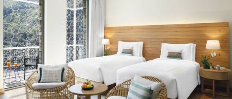 Quarto de hotel com duas camas, cadeiras de vime, uma mesa com frutas e uma varanda com vista para o verde.