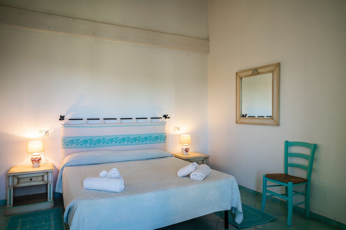 Quarto de hotel com uma cama, duas mesas de cabeceira com luminárias, uma cadeira e um espelho na parede.