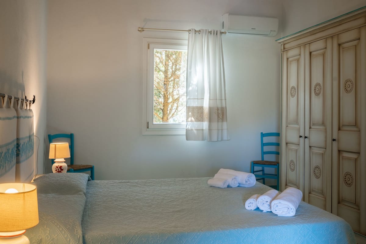 Quarto com cama, mesa de cabeceira com abajur, janela com cortinas e guarda-roupa.