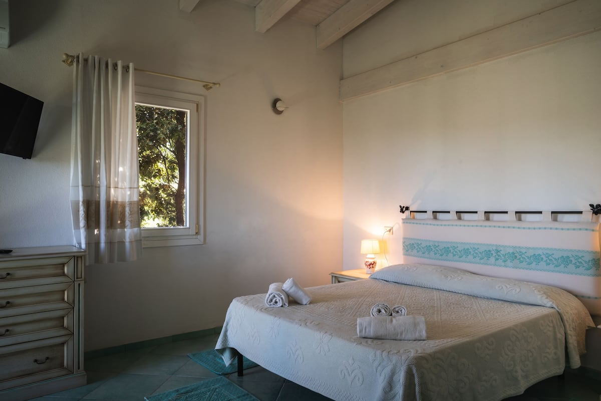 Quarto com cama, mesa de cabeceira, luminária, TV na parede e janela com cortinas.